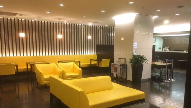Um lobby com sofás e mesas amarelas, uma recepção e uma planta em vaso grande.