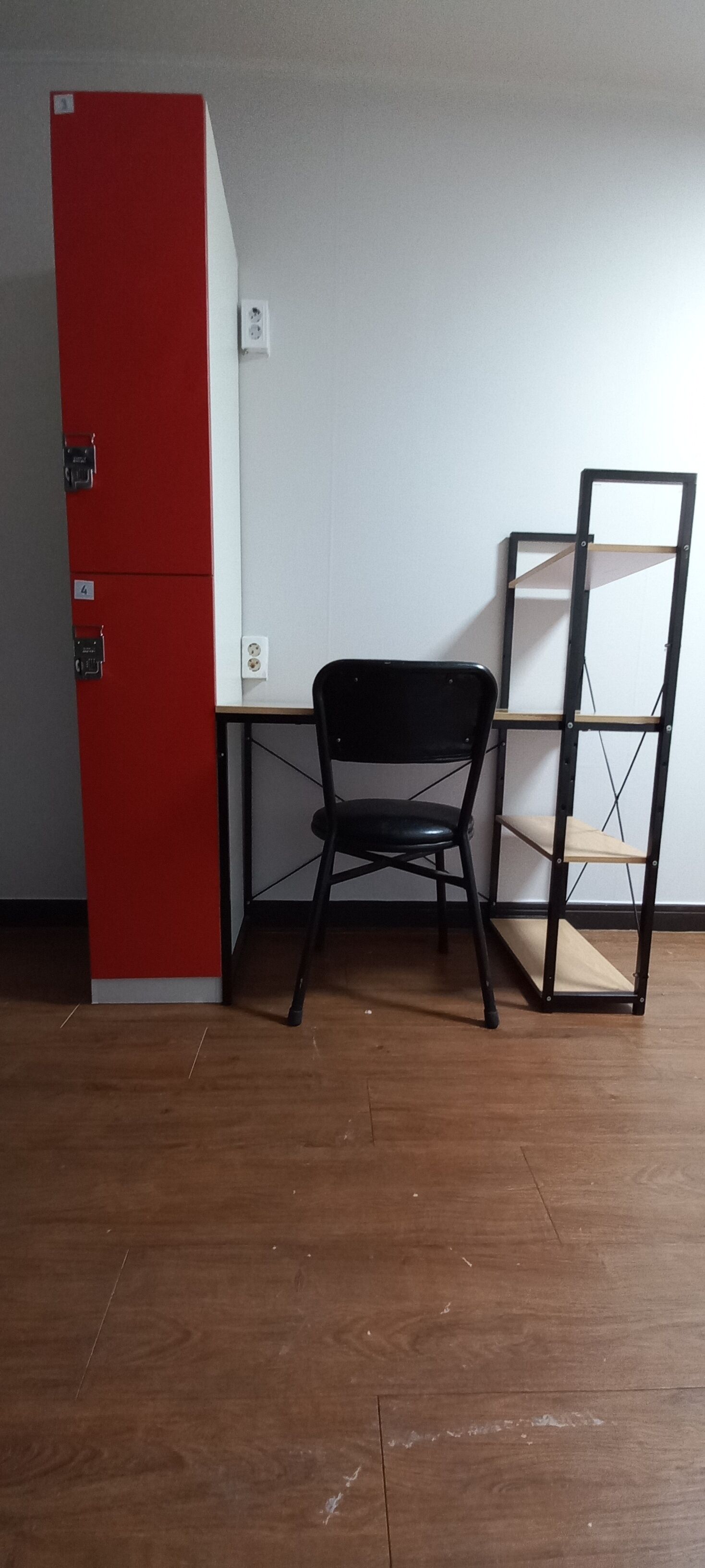 Quarto com piso de madeira, uma cadeira preta, um armário vermelho e uma estante de metal na parede.