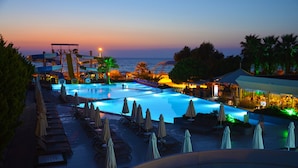 Uma área de piscina em resort, com espreguiçadeiras e guarda-sóis, um toboágua e vista para o oceano ao entardecer.