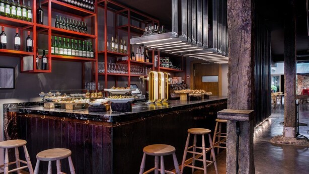 Uma área de bar com bancada de mármore, banquetes, prateleiras com garrafas e um grande pilar de madeira.