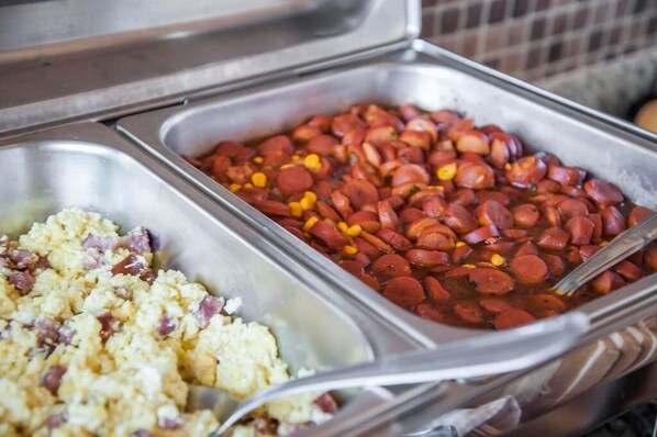 Buffet com diversas opções de comida, incluindo salsichas, ovos mexidos e um prato com milho e bacon.