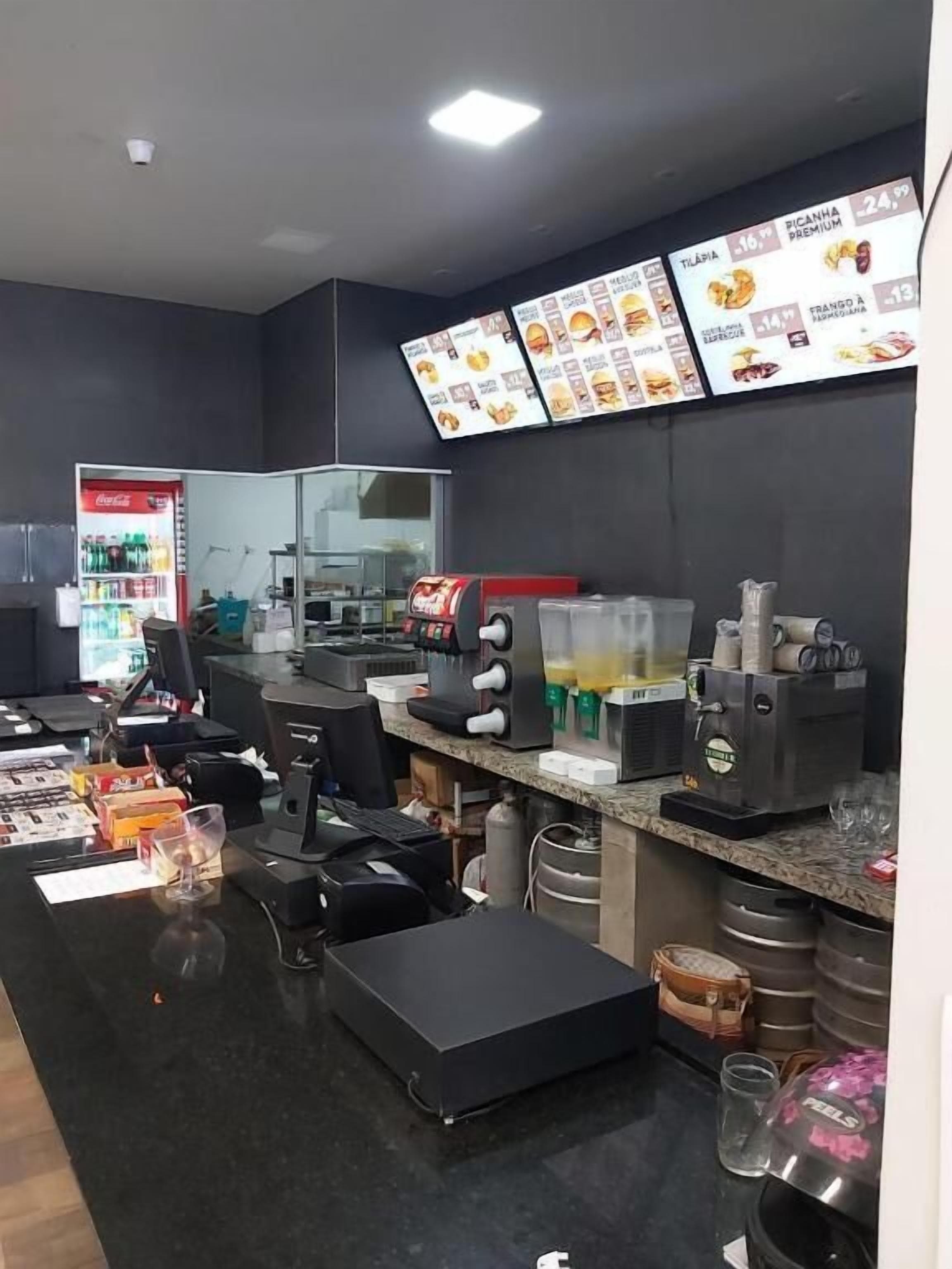 Interior de um restaurante de fast-food, com uma estação de bebidas de serviço próprio, um refrigerador de Coca-Cola e um quadro de menu exibindo os pratos.