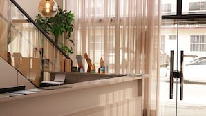 Uma receção moderna com um computador portátil, papéis e objetos decorativos. Uma planta em vaso é visível à esquerda, e uma porta de vidro dá para o exterior.