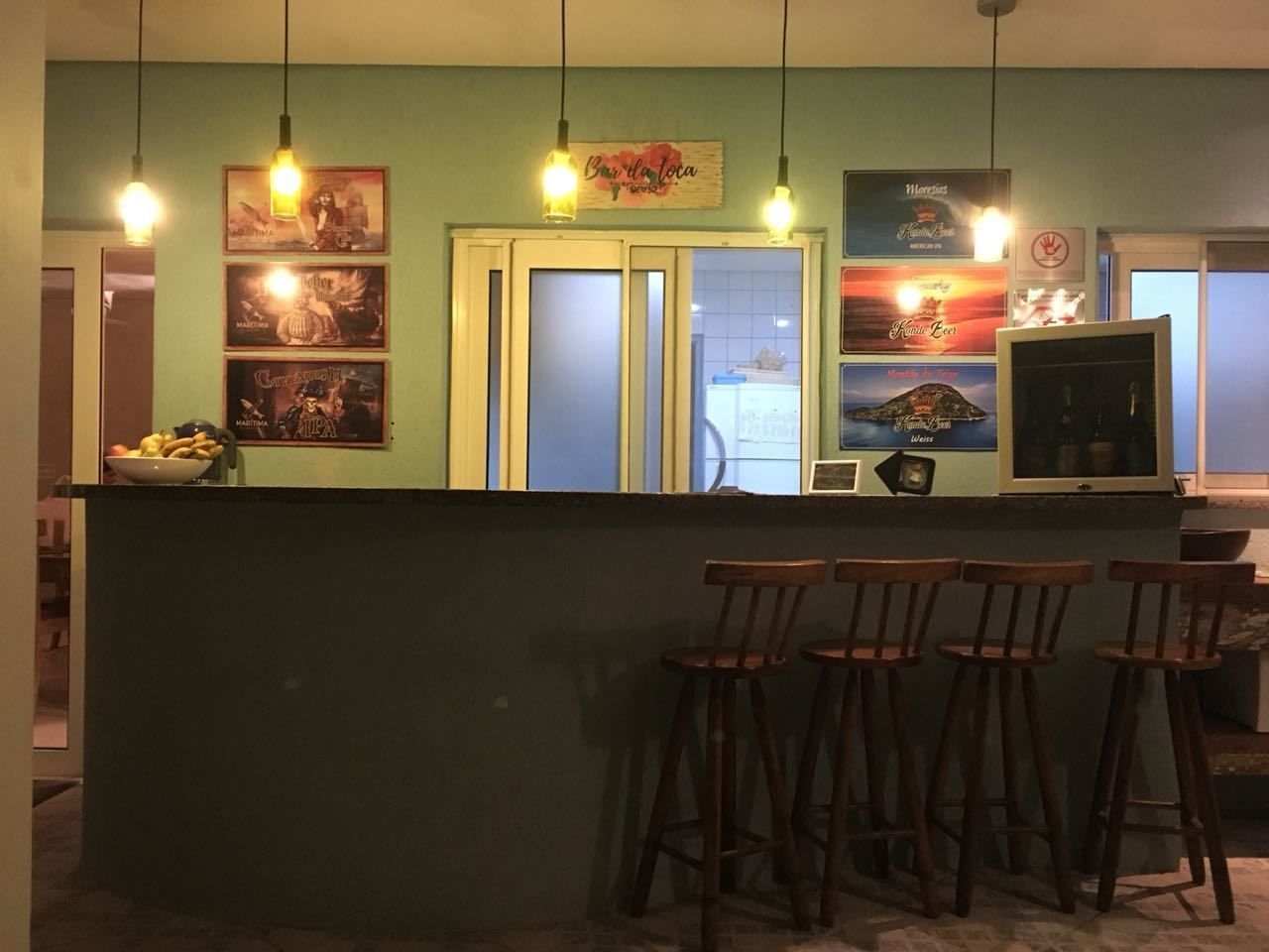 Uma área de bar com balcão e banquetas, cartazes emoldurados na parede e luminárias pendentes.
