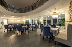 Um restaurante espaçoso com teto curvo e varanda. Mesas dispostas em formato de semi-círculo, com plantas em vasos e lustres.