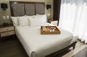 Cama bem arrumada com lençóis brancos, mesa de cabeceira de madeira com telefone e abajur, e uma bandeja com itens de café da manhã sobre a cama.
