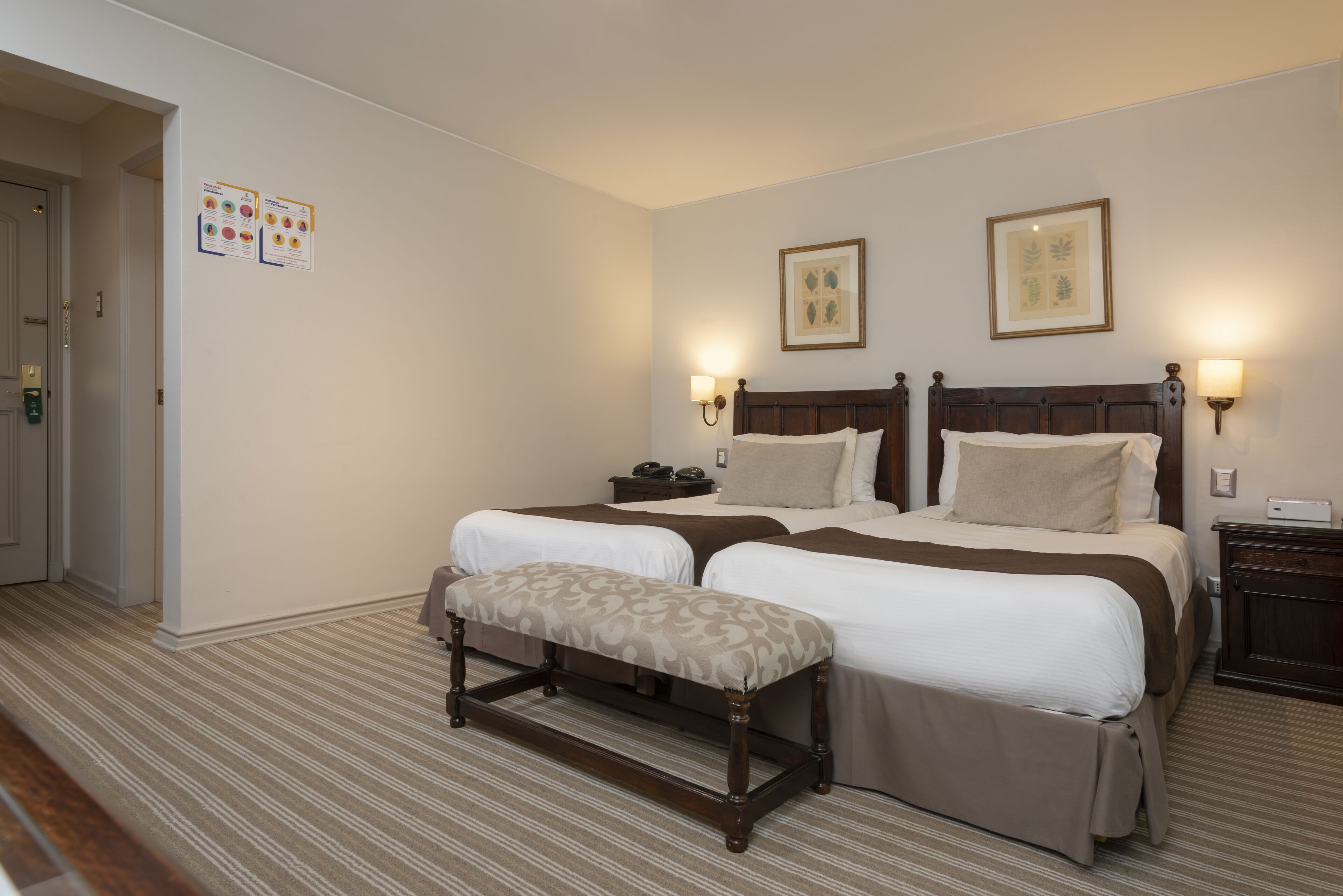 Quarto de hotel com duas camas, um banquinho e uma mesa de cabeceira com abajur.