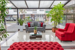 Um espaço moderno e interno com um sofá cinzento, poltrona vermelha e um banquinho estofado vermelho. Há plantas em vasos e uma mesa de centro de vidro.