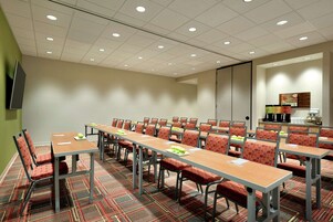 Sala de conferências com várias mesas e cadeiras, equipada com uma televisão de tela plana e um espaço para café.