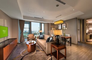 Quarto de hotel moderno com uma grande janela que oferece vista da cidade, um sofá, uma mesa de jantar com cadeiras, uma TV e uma pequena mesinha lateral.