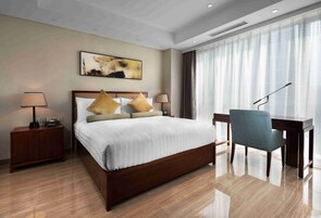 Quarto de hotel com uma cama grande, duas mesas de cabeceira com abajures, uma escrivaninha com cadeira e uma janela ampla com cortinas.