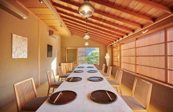 Uma sala de jantar tradicional japonesa com uma mesa comprida posta para quatro pessoas, vigas de madeira no teto e portas deslizantes à direita.