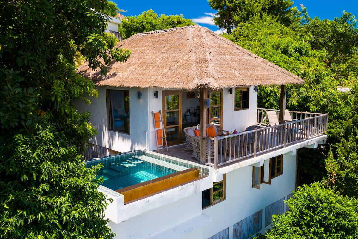 Uma casa de dois andares com telhado de colmo, piscina e varanda com mobiliário de exterior.