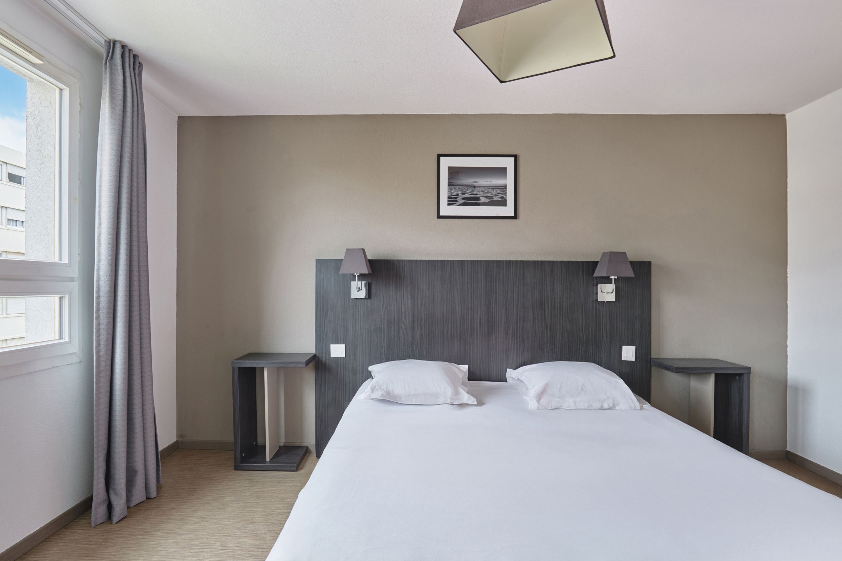 Quarto de hotel com uma cama grande, duas mesas de cabeceira, cabeceira, janela com cortinas e um quadro na parede.