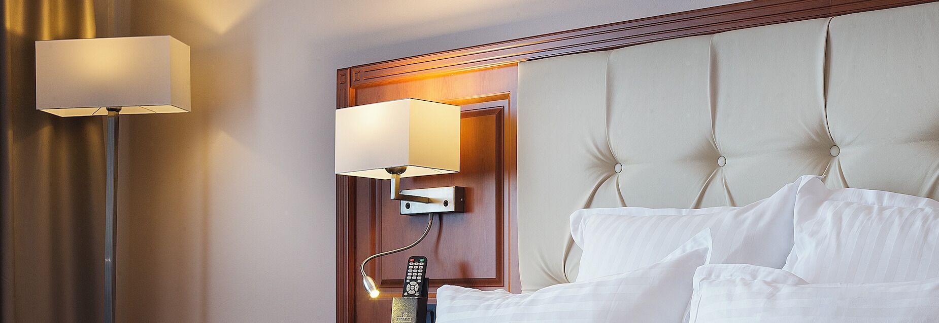 Quarto de hotel com cabeceira estofada, abajur e controle remoto na mesa de cabeceira.