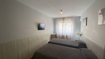 Quarto com duas camas, TV fixada na parede, janela com cortinas florais e um quadro na parede.
