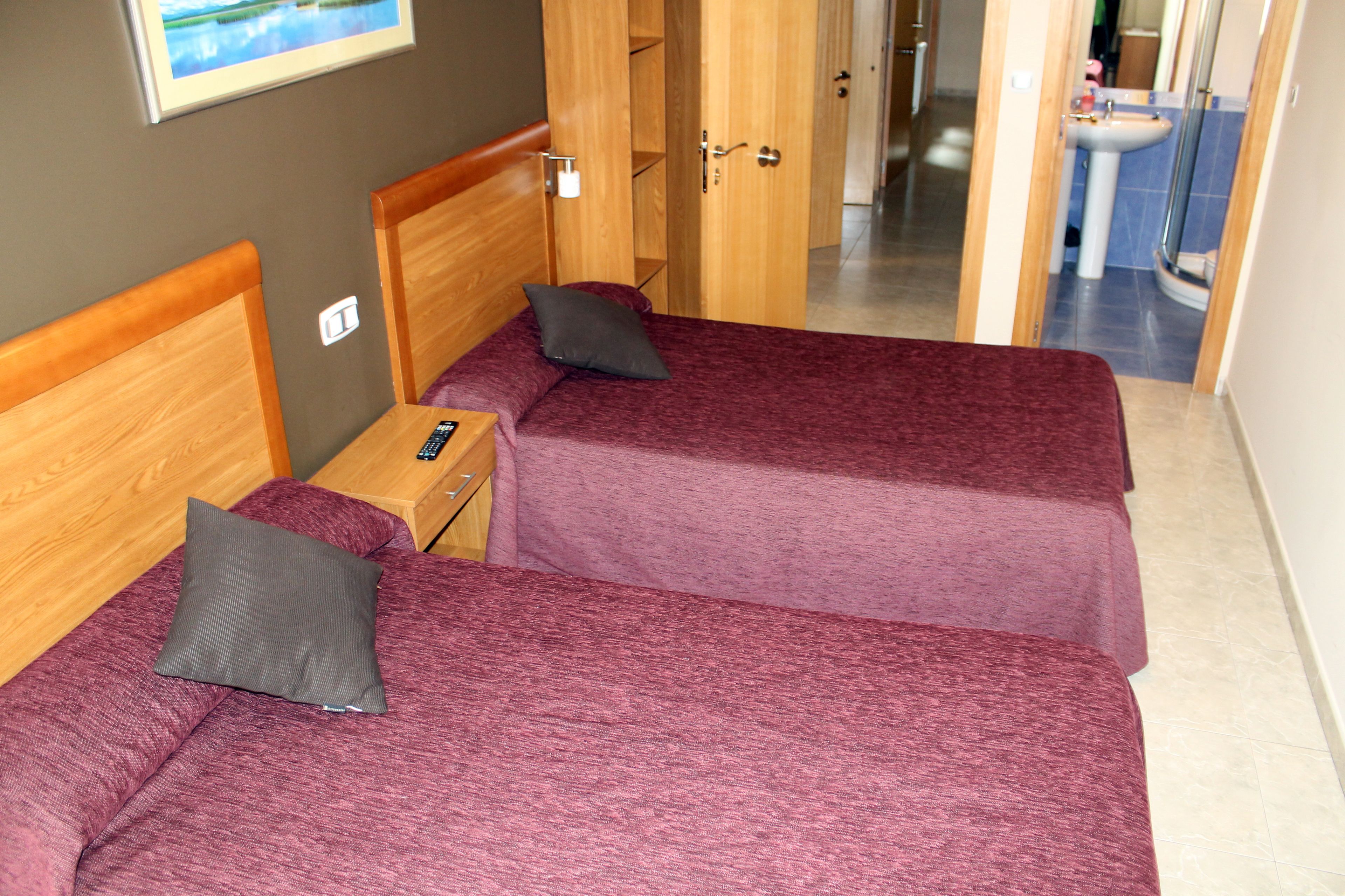 Quarto de hotel com duas camas, cada uma com cabeceira de madeira e colcha vermelha escura. Há uma pequena mesa de cabeceira de madeira com um controle remoto.