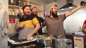 Cozinha com eletrodomésticos de aço inoxidável, pia e fogão. Há panelas e frigideiras na bancada, e uma pessoa está cozinhando.