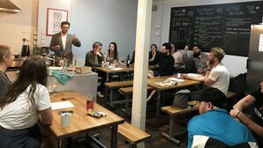 Um grupo de pessoas em uma sala moderna e bem iluminada, com mesas e bancos de madeira, uma lousa com informações sobre eventos e um aviso sobre a história do museu.