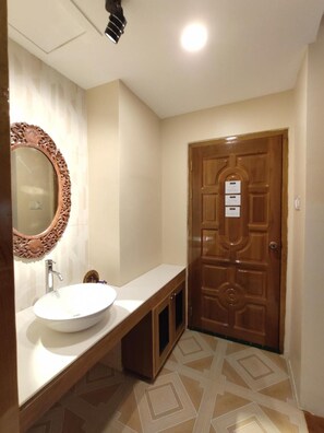 Um banheiro com pia branca, toucador de madeira, espelho com moldura ornamentada e porta de madeira com uma placa.