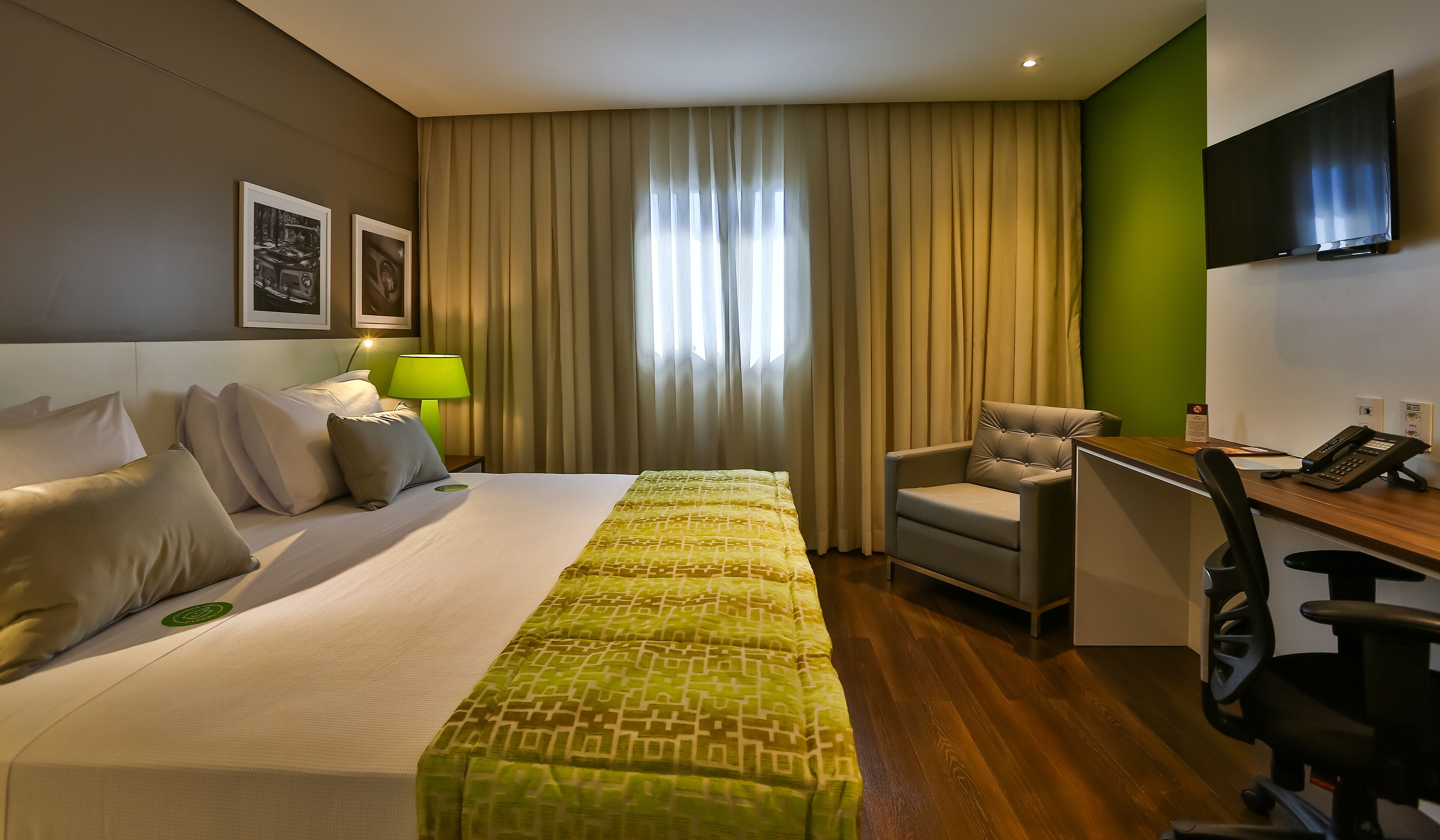 Quarto de hotel com uma cama, uma escrivaninha com telefone, uma cadeira e uma televisão na parede.