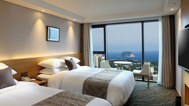 Quarto de hotel com duas camas, uma mesa de escritório, televisão e uma janela grande com vista para a cidade.
