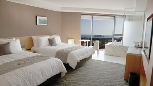 Quarto de hotel com duas camas, uma janela ampla com vista, uma pequena mesa e área de banheiro.