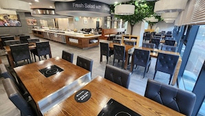 Um restaurante moderno com mesas de madeira e cadeiras pretas, um balcão central com a placa que diz “Salada Fresca com Bem-Estar”, e vista para a rua através de grandes janelas.