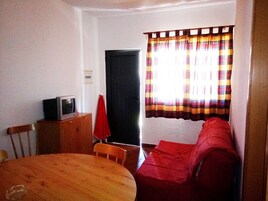 Uma sala de estar com uma mesa de madeira, um sofá vermelho, uma televisão sobre um móvel de madeira e uma janela com cortina xadrez.