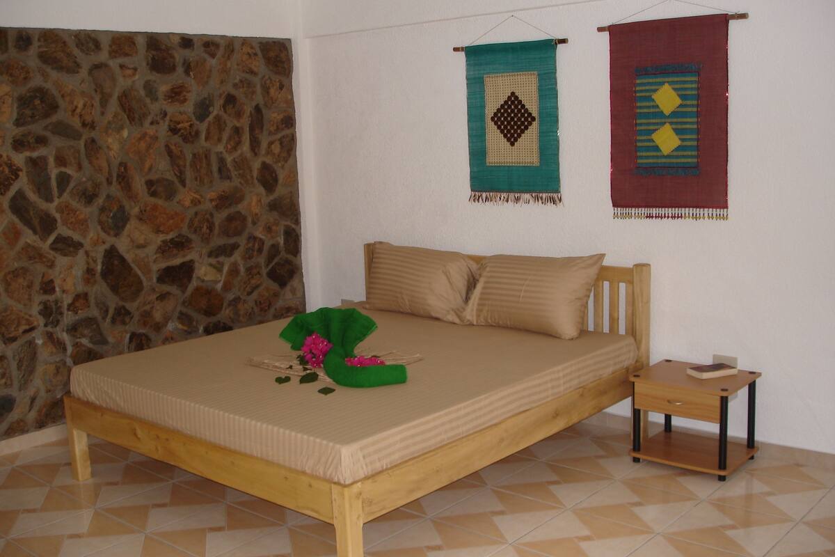 Uma cama com estrutura de madeira, uma pequena mesa de cabeceira e uma parede de pedra.