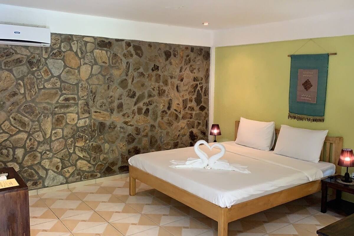 Quarto de hotel com parede de pedra, cama com roupa branca, cabeceira de madeira e mesa de cabeceira com abajur.