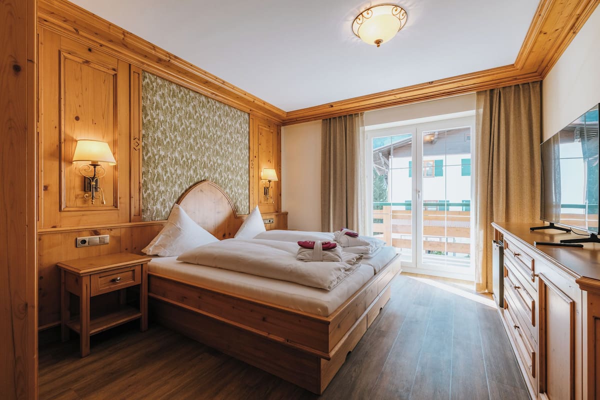 Quarto com estrutura de madeira, cama, luminárias de cabeceira, cômoda e vista através de uma porta de vidro.