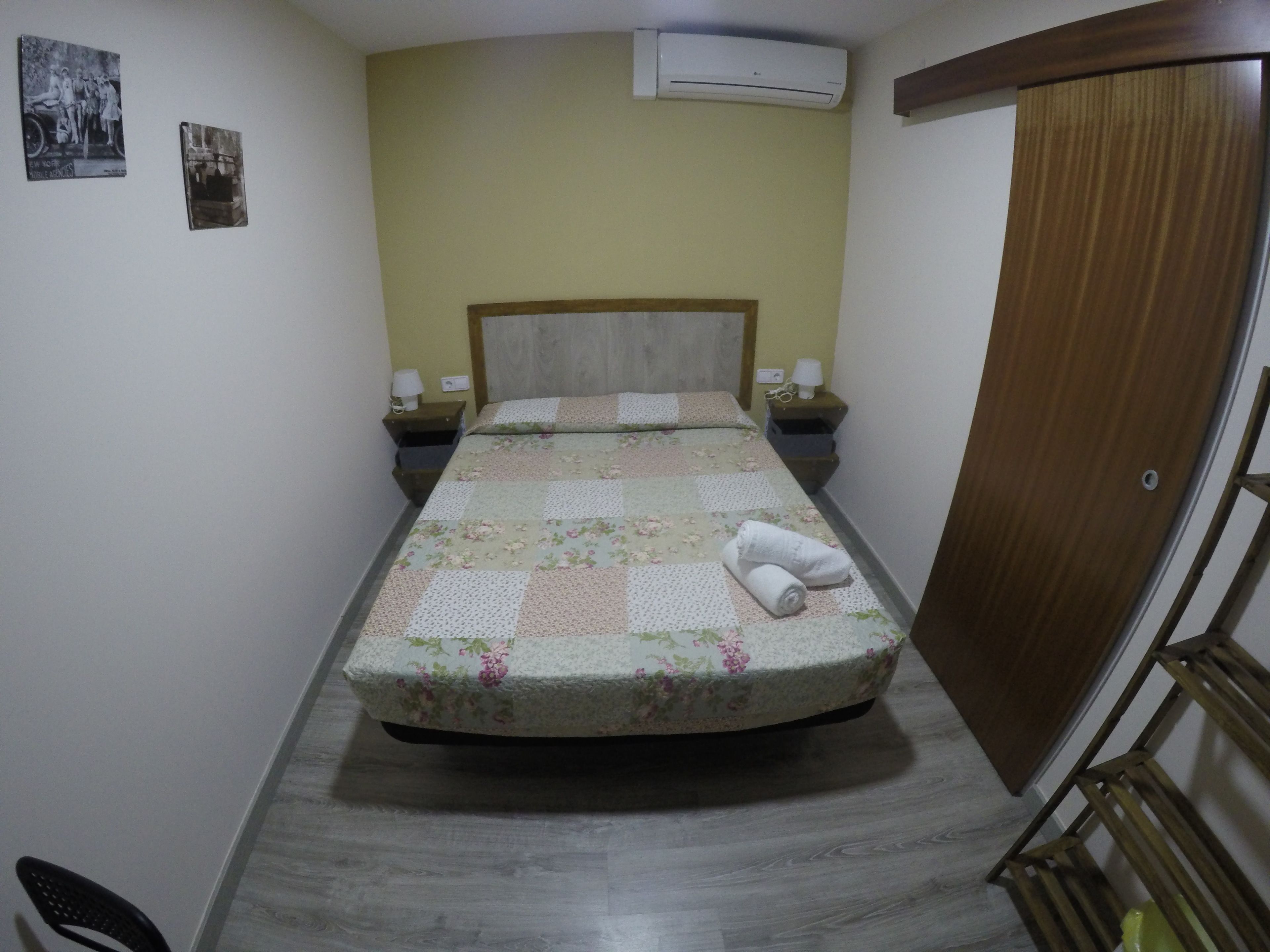 Quarto pequeno e bem arrumado, com cama, mesas de cabeceira, um ar condicionado e porta de madeira.