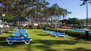 Área à beira da piscina com espreguiçadeiras e guarda-sóis, cercada por árvores e um céu claro.