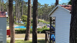 Uma pequena cabana branca com varanda, rodeada por árvores e uma área de piscina ao fundo.