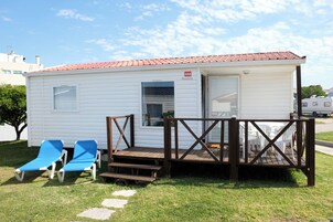 Uma casa móvel branca pequena com teto vermelho, um alpendre com grades de madeira e duas cadeiras de jardim azuis.