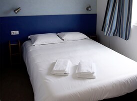 Cama bem arrumada com lençóis brancos e duas toalhas dobradas, cabeceira azul e luminária fixada na parede.