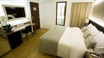 Quarto de hotel com cama, escrivaninha, cadeira, televisão e banheiro visível por uma porta aberta.