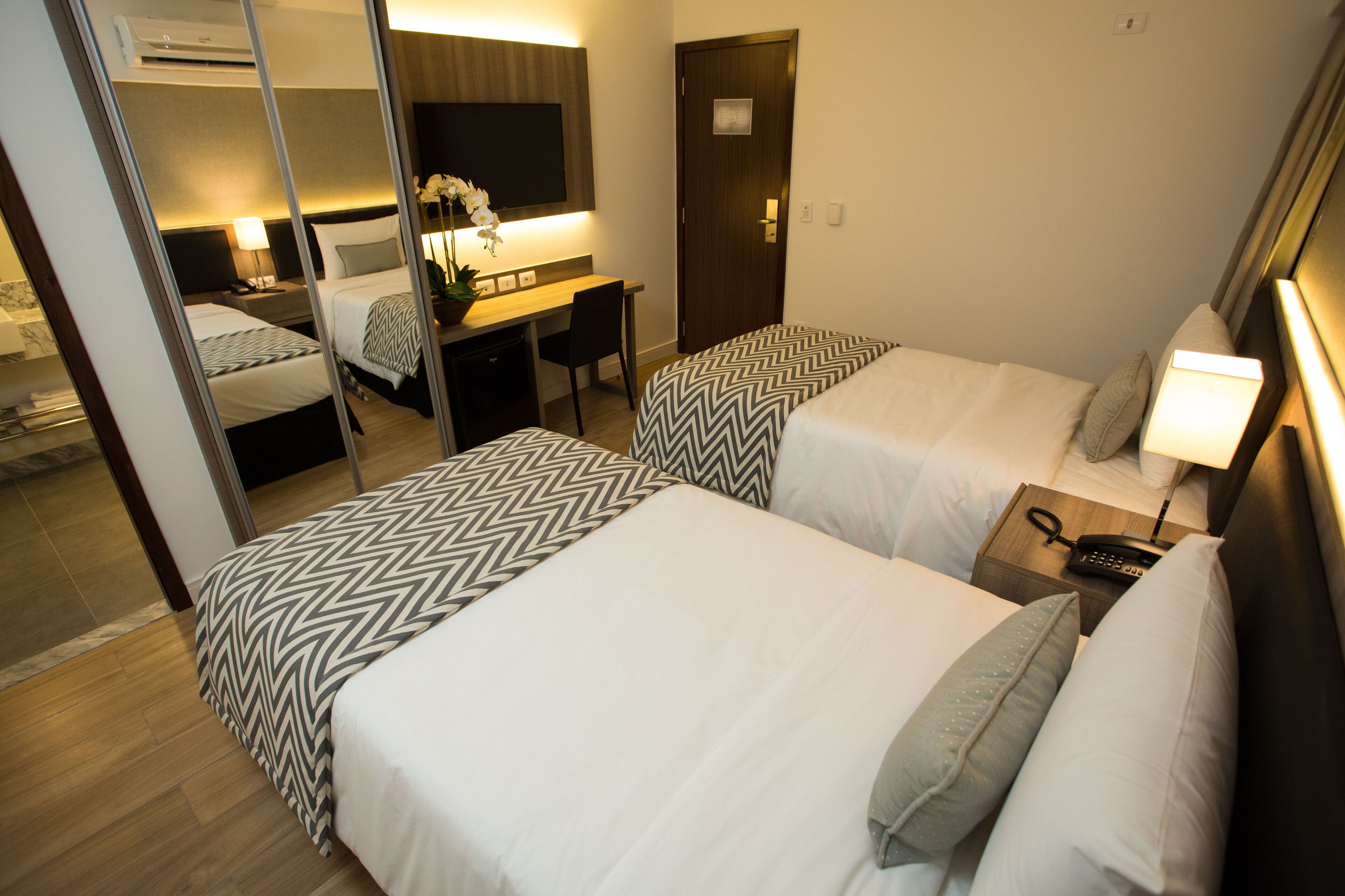 Quarto de hotel com duas camas, uma escrivaninha, uma cadeira e um espelho.
