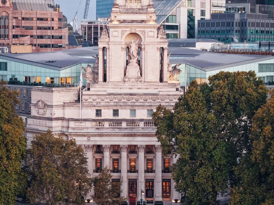 Um edifício clássico com uma estátua central, rodeado por um jardim ajardinado e arranha-céus modernos ao fundo.