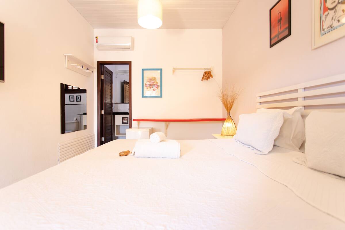 Quarto de hotel com uma cama grande, uma mesinha de cabeceira com abajur, um quadro na parede e uma pequena janela.