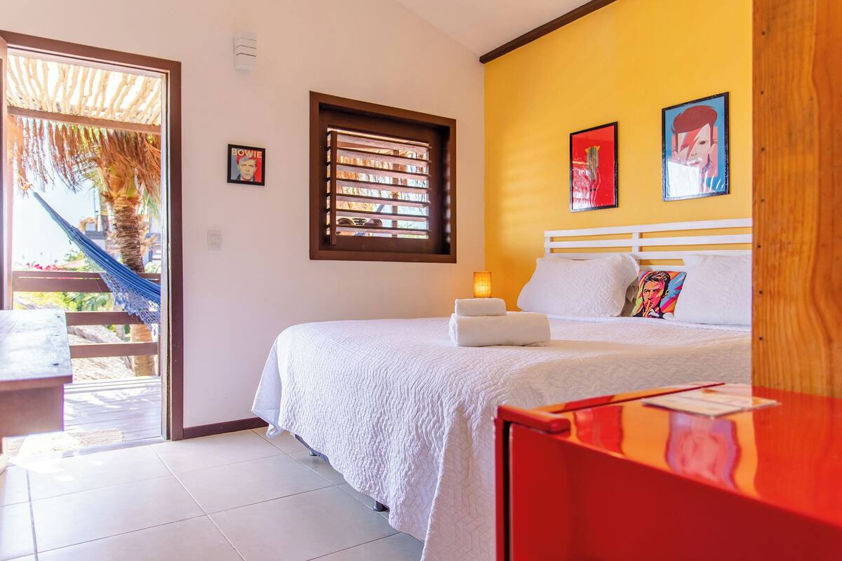 Quarto com cama, janela com persianas, porta que dá para área externa e parede com quadros.