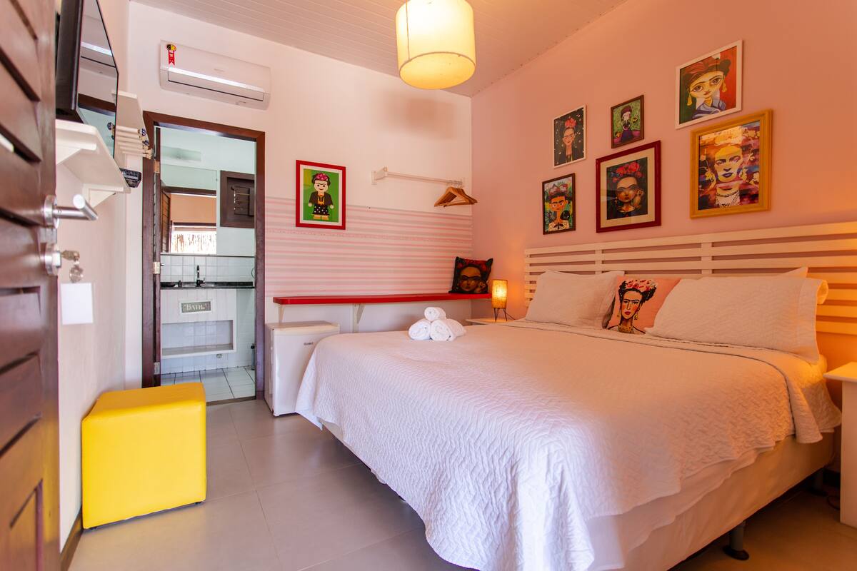 Quarto de hotel com cama, um banquinho amarelo pequeno, banheiro com pia e espelho, e obras de arte nas paredes.