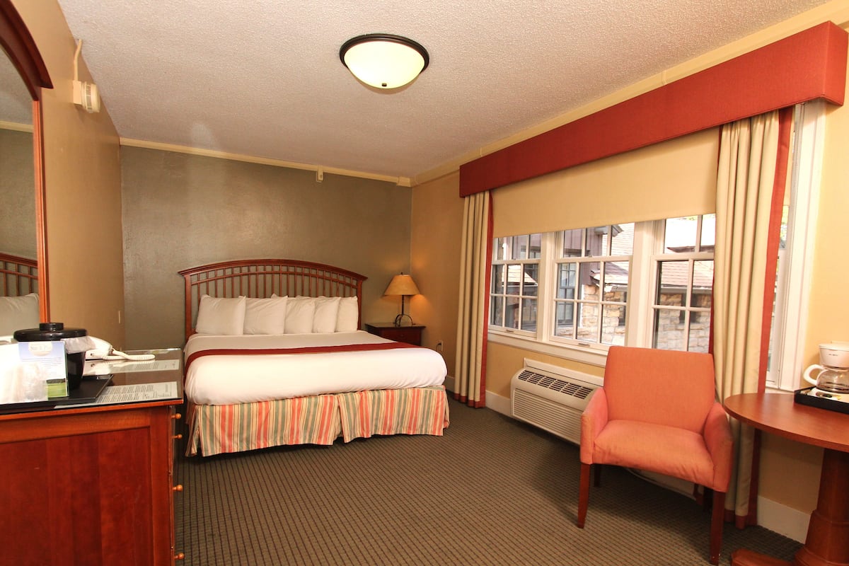 Quarto de hotel com cama, escrivaninha com telefone e luminária, cadeira e janela com cortinas.