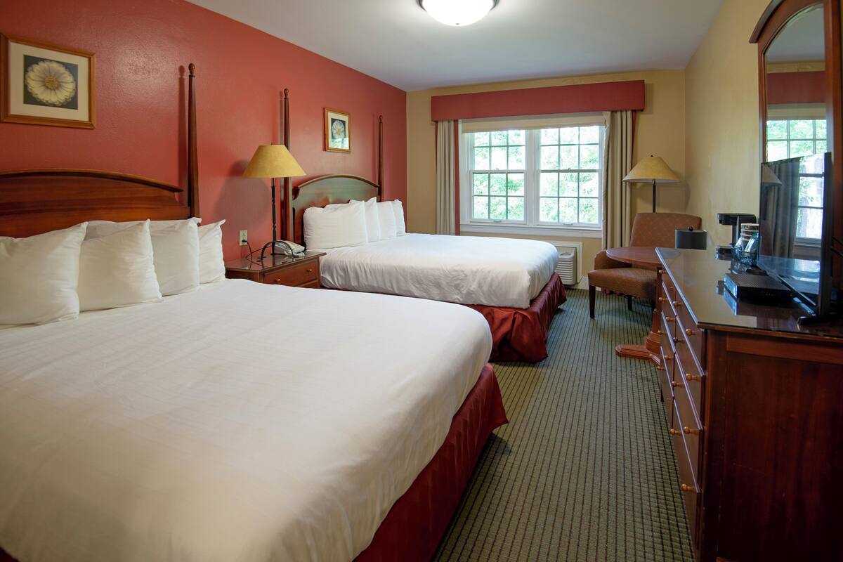Quarto de hotel com duas camas, uma escrivaninha, uma cadeira, cômoda e janela com cortinas.