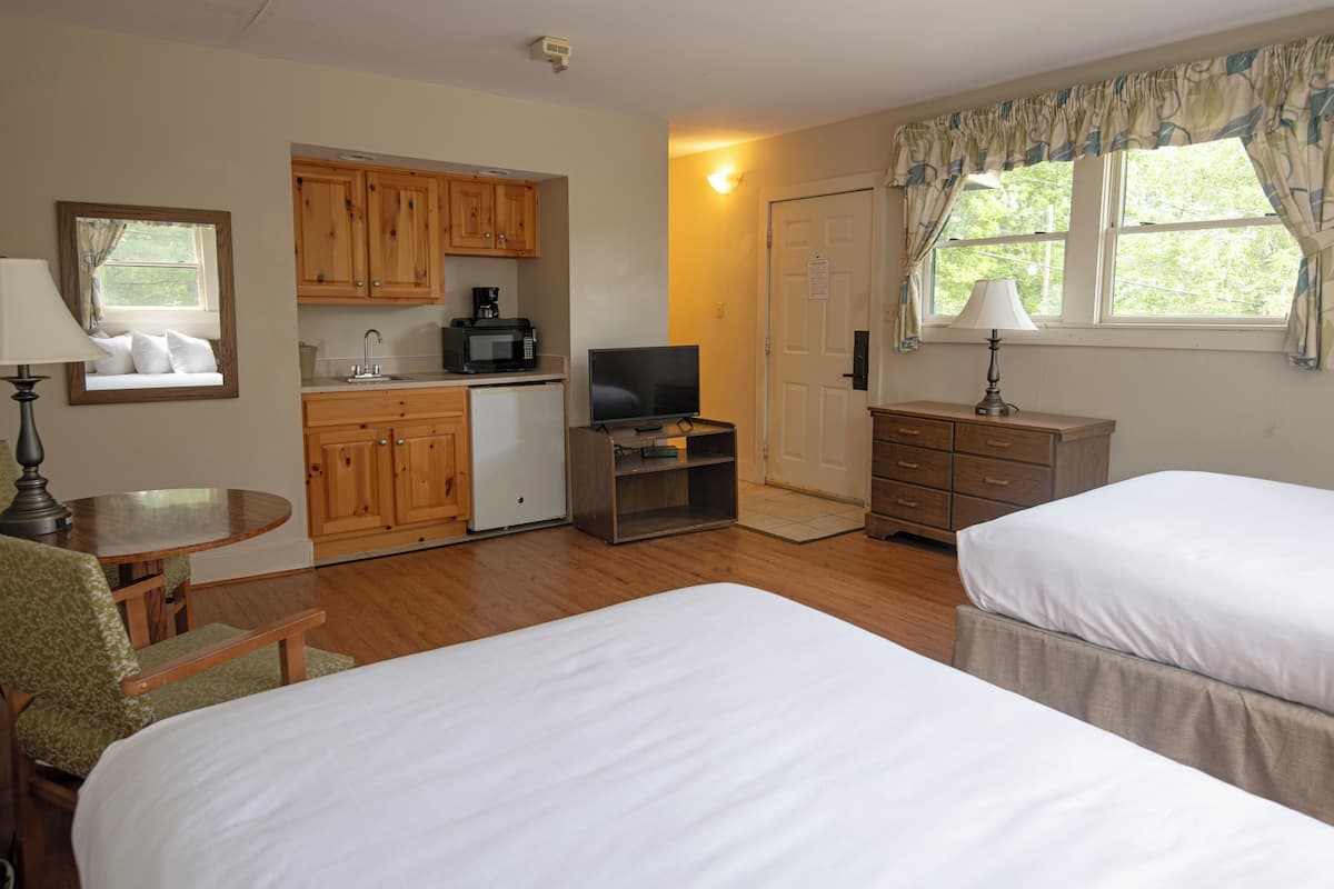 Quarto de hotel com duas camas, uma pequena cozinha compacta, televisão e uma mesa de trabalho com cadeira.