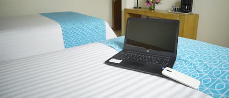 Quarto de hotel com cama, secretária, laptop, máquina de café e televisão na parede.