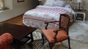 Quarto com parede de pedra, cama com roupa de cama listrada, cadeira de madeira, mesa pequena e abajur em um criado-mudo.