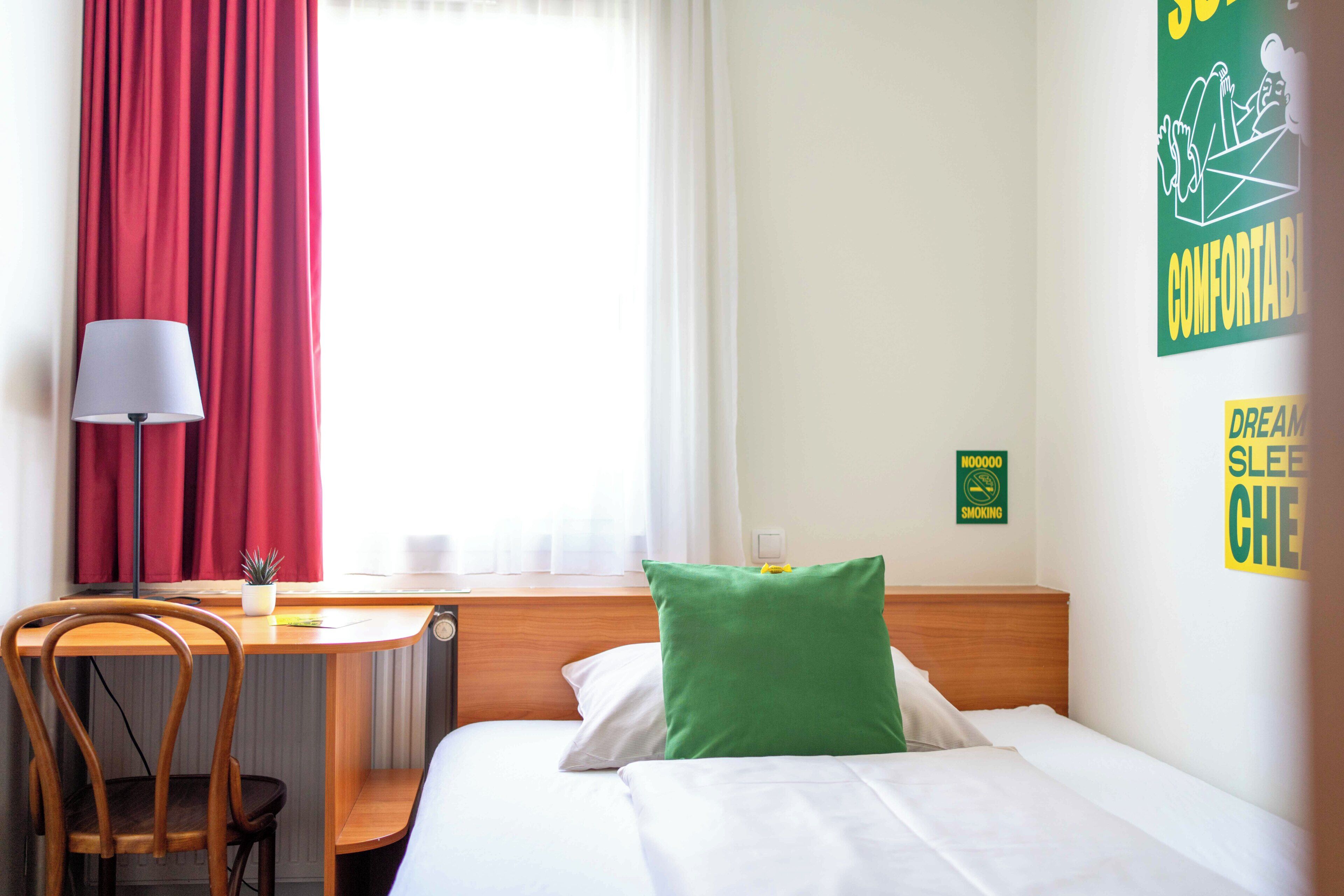 Quarto de hotel com cama, escrivaninha, cadeira, luminária e janela com cortinas.