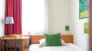Quarto de hotel com cama, escrivaninha, cadeira, luminária e janela com cortinas.